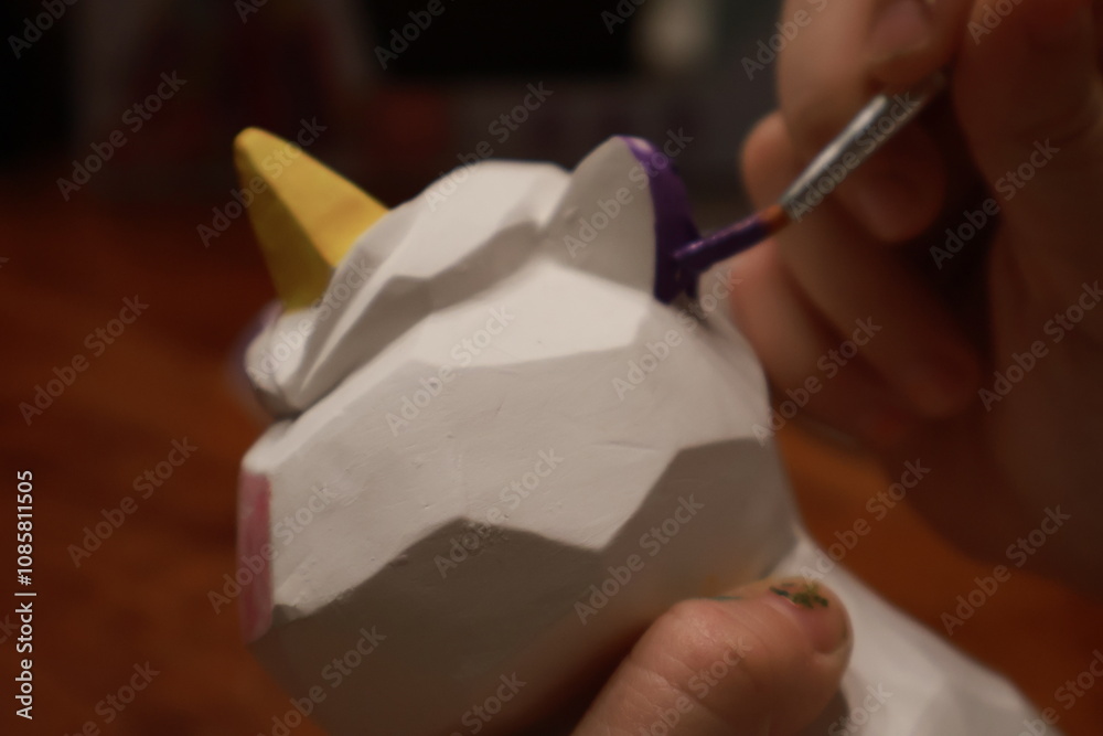 The photo shows a child painting a unicorn figurine, focused on adding ...