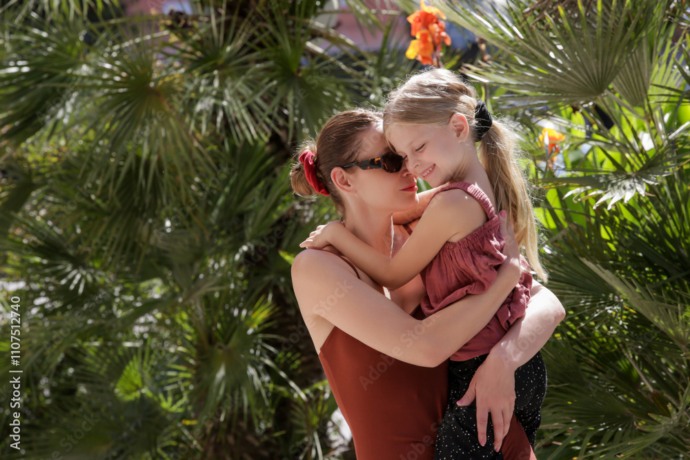 Candid portrait of a mother and daughter hugging in the street with beautiful greenery behind ...