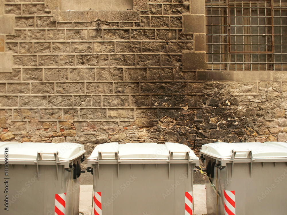 Three gray garbage bins lined up against an old stone wall with a metal ...