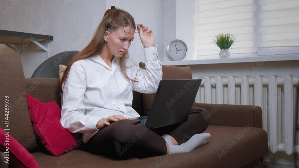 Young woman adjusting her glasses while frowning at laptop screen ...