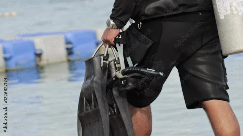 Dive instructor in wet suit carries his gear oxygen tank and fins ...