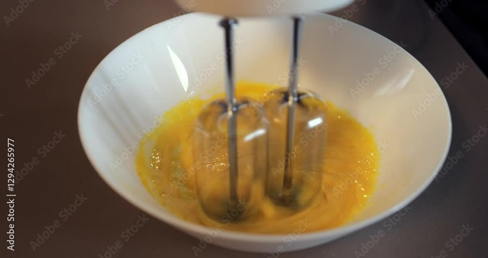 Electric mixer whips egg yolks in a white deep bowl, creating a smooth ...
