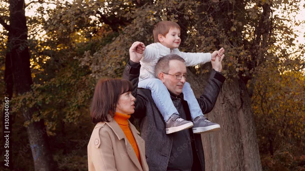 Father carries son on shoulders enjoying serene stroll through park near wife. Father carries ...