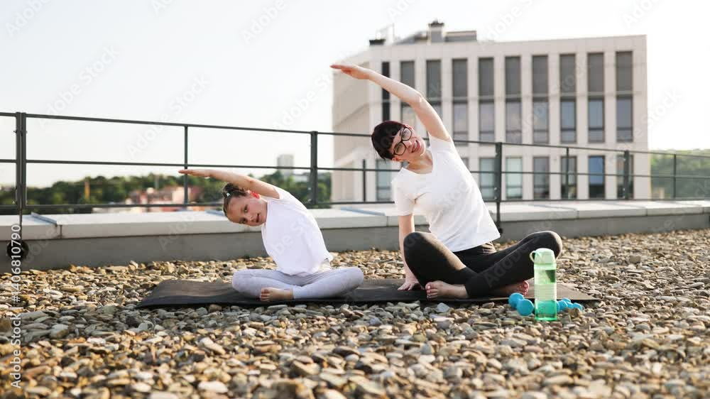 Mother and daughter sitting on yoga mats stretching on rooftop during ...