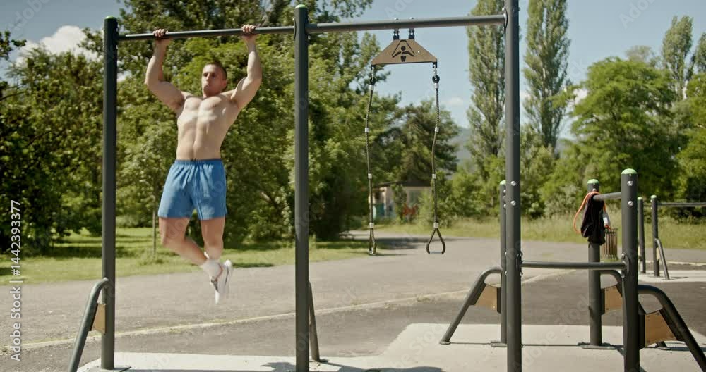 A muscular man performs pull-ups on a pull-up bar in a park on a sunny ...